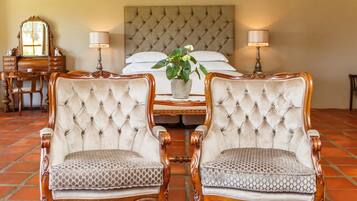 Quarto de hotel com uma cama, duas poltronas estofadas com almofadas estampadas, uma mesinha de cabeceira com abajur, cômoda e um vaso de planta sobre a cama.