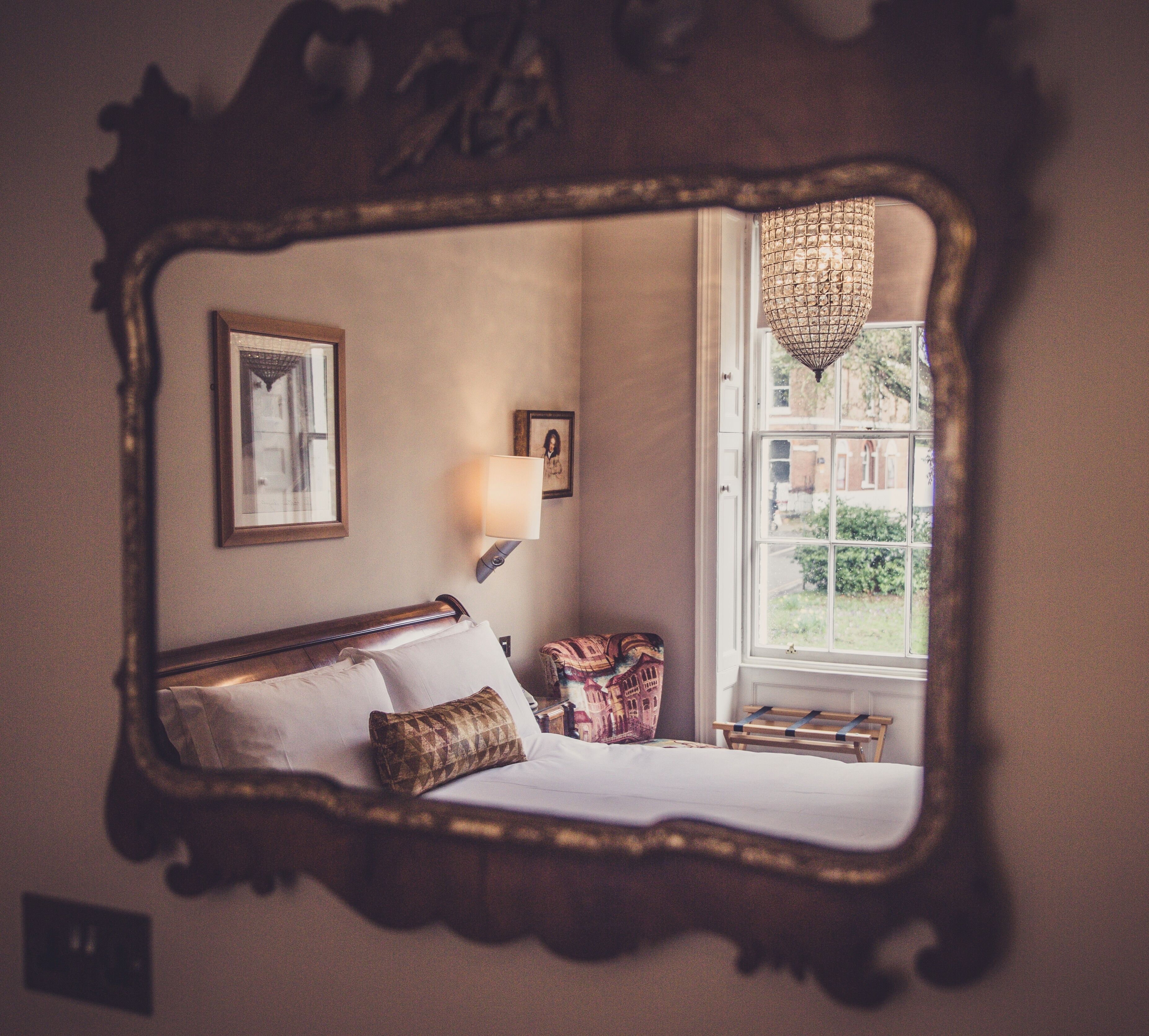 Quarto com um espelho grande refletindo uma cama, uma janela com vista para vegetação e um candelabro.