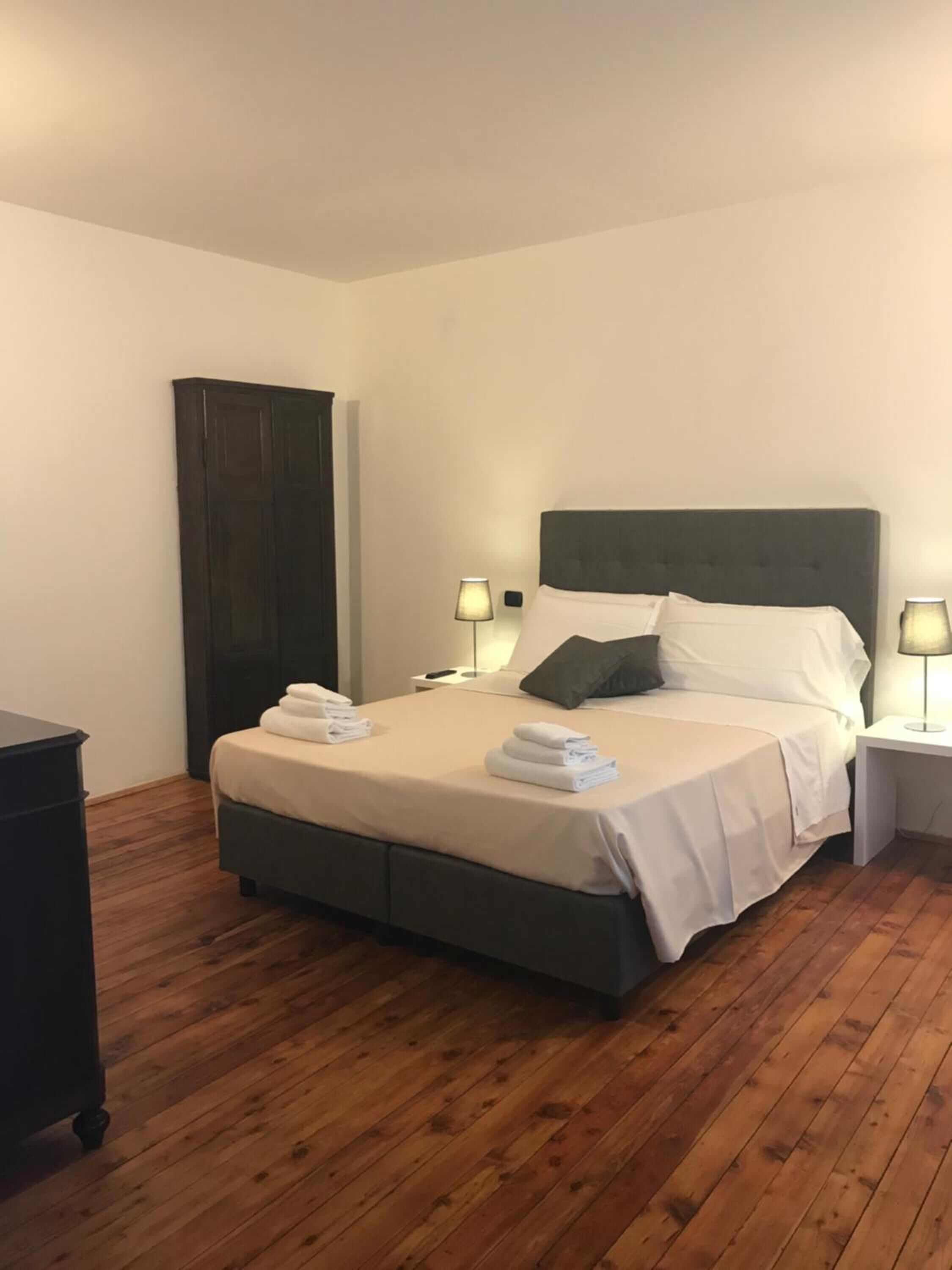 Cama bem arrumada com lençóis brancos e cabeceira escura, duas luminárias de apoio e um guarda-roupa escuro em um quarto com piso de madeira.