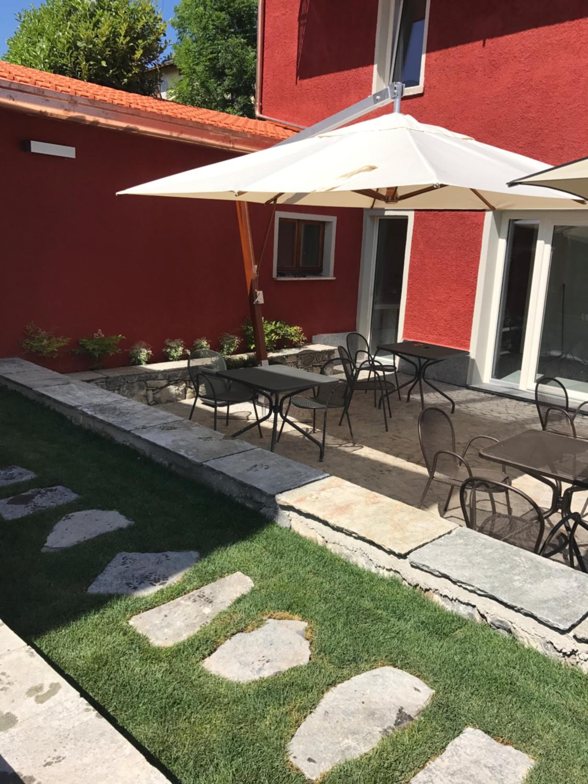 Um pátio com mesas ao ar livre, um guarda-sol branco e um edifício vermelho ao fundo.
