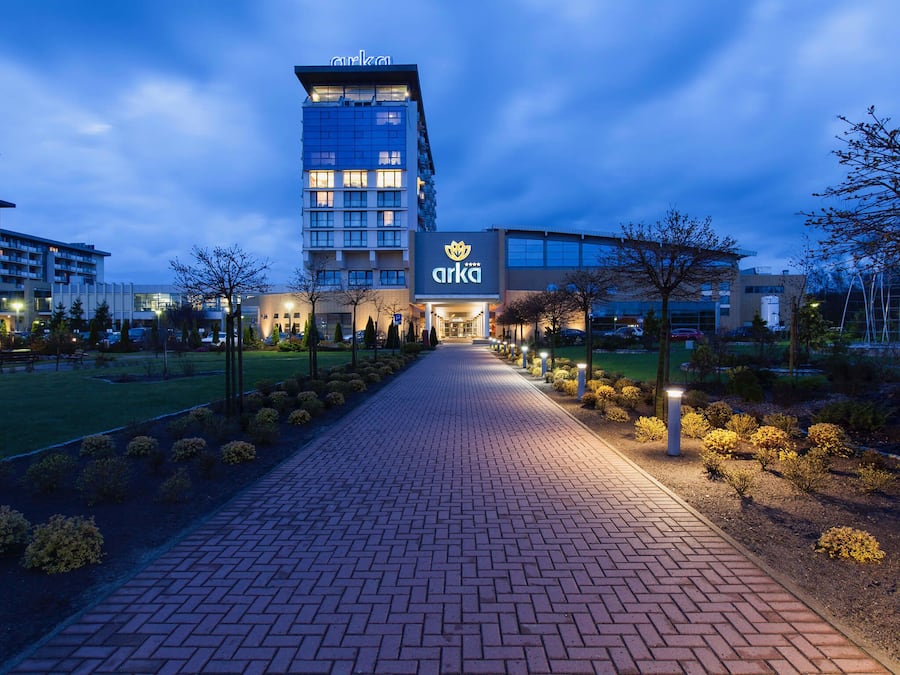 Entrada de um hotel com um caminho pavimentado que leva ao edifício, cercada por áreas ajardinadas e árvores.