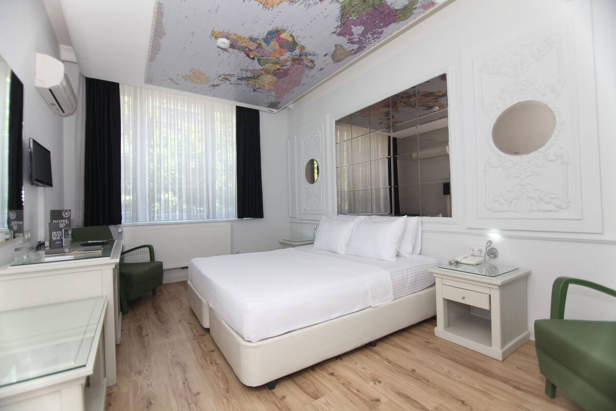 Quarto de hotel com cama, escrivaninha, cadeira, mesa de cabeceira, janela com cortinas, televisão na parede e teto com mapa.