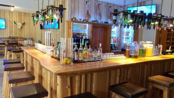 Uma área de bar com banquetas de madeira, um balcão com diversos garrafas e copos, e uma televisão presa à parede.
