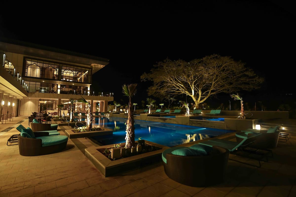 Área de piscina à noite com espreguiçadeiras e uma grande árvore.
