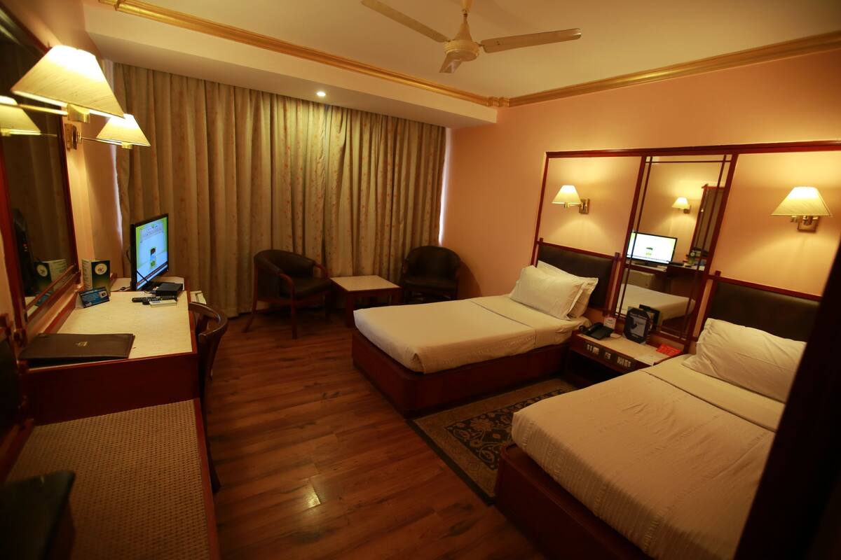 Quarto de hotel com duas camas, uma mesa de escritório com computador, uma cadeira, luminária de mesa, ventilador de teto e janela com cortinas.