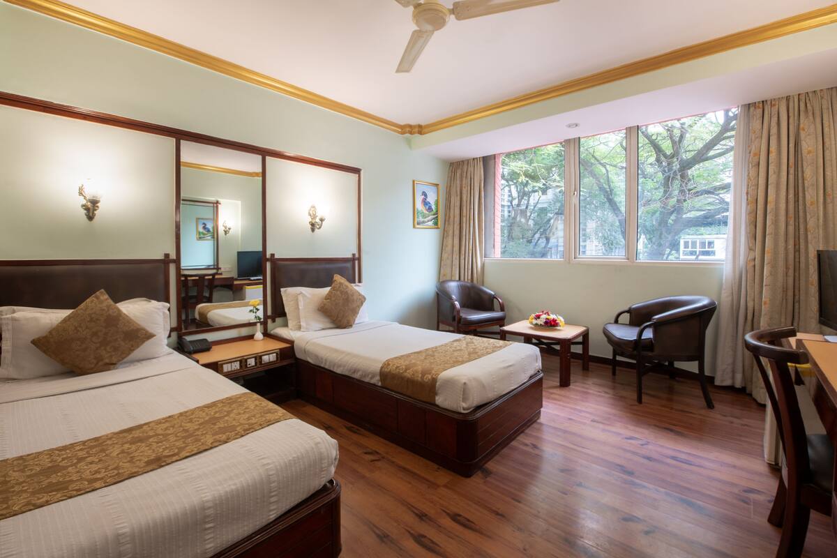 Quarto de hotel com duas camas, uma escrivaninha, uma cadeira, televisão e janela com vista para árvores.
