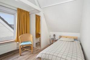 Quarto com uma cama, uma cadeira, uma mesinha e uma janela com cortinas.