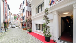 Rua de paralelepípedos que conduz a um edifício com entrada de tapete vermelho, plantas em vasos e uma bandeira sobre a porta.