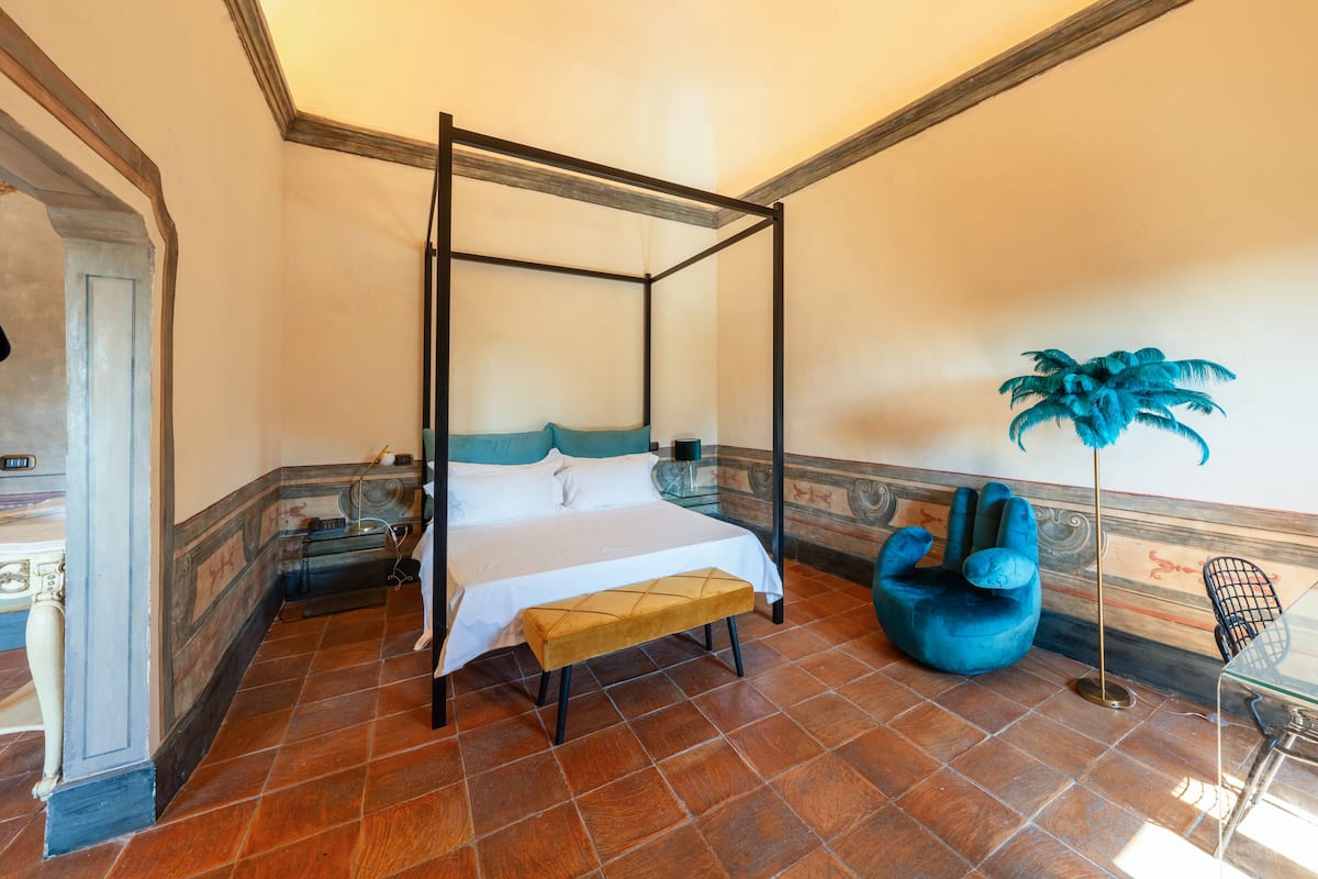 Uma cama com dossel, um banco, uma cadeira azul e uma planta alta em um quarto com piso de azulejos e paredes decorativas.