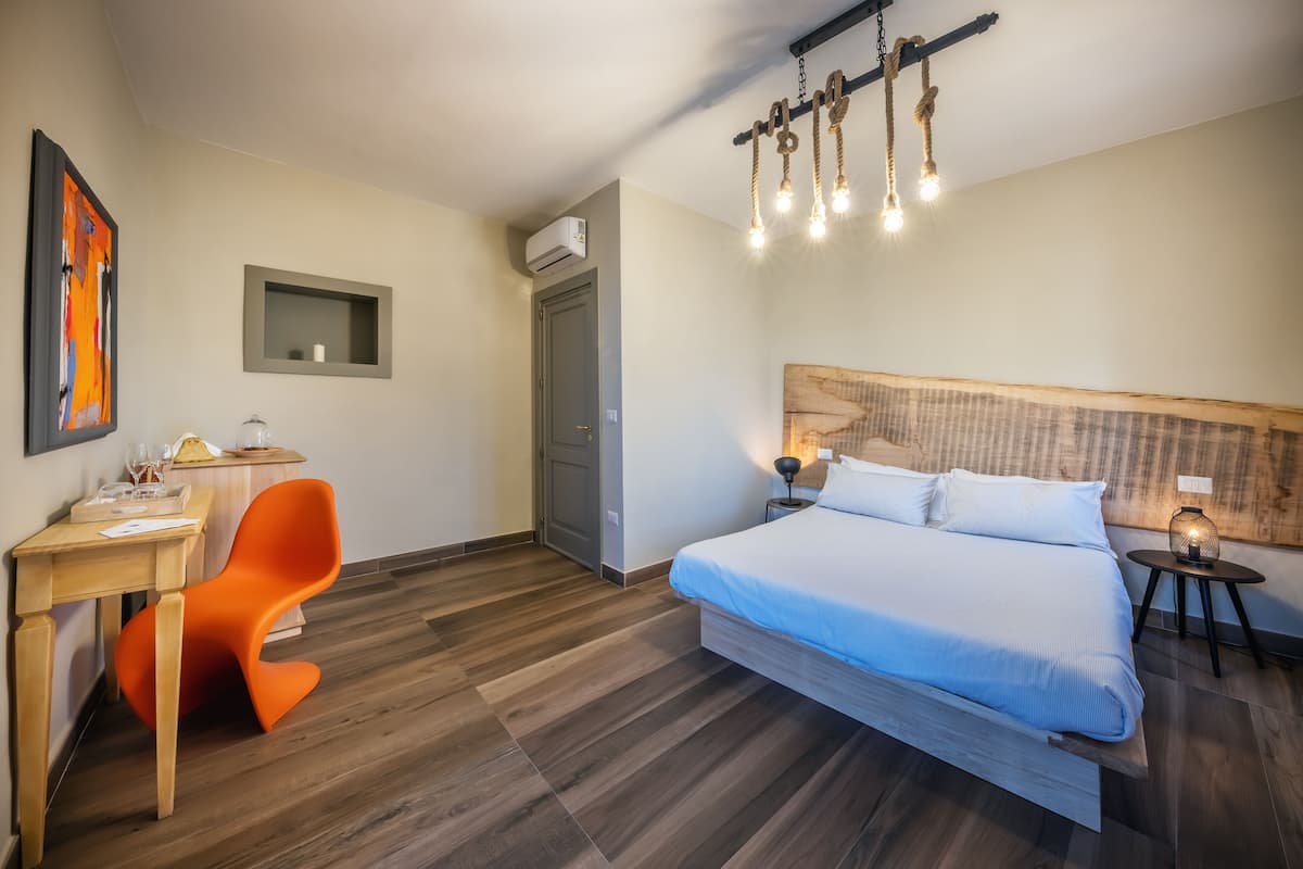 Quarto de hotel moderno com piso de madeira, cama com cabeceira, escrivaninha com cadeira, mesinha com luminária e ar-condicionado fixado na parede.
