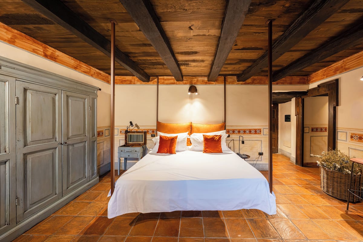 Quarto com cama grande, vigas de madeira no teto e piso ladrilhado.