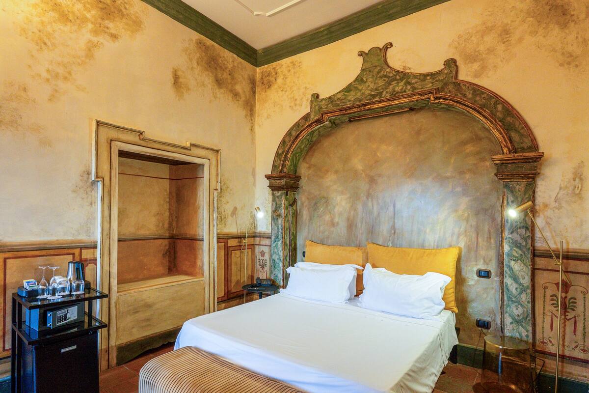 Quarto de hotel com uma cama grande, uma mesa de cabeceira com abajur e um minibar.