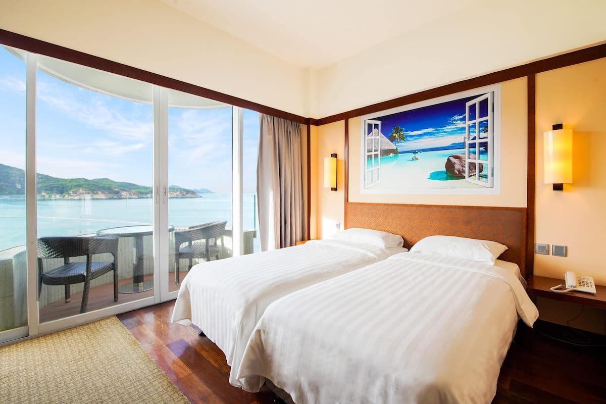Quarto de hotel com duas camas, varanda com vista para o mar e uma pintura de uma paisagem tropical.