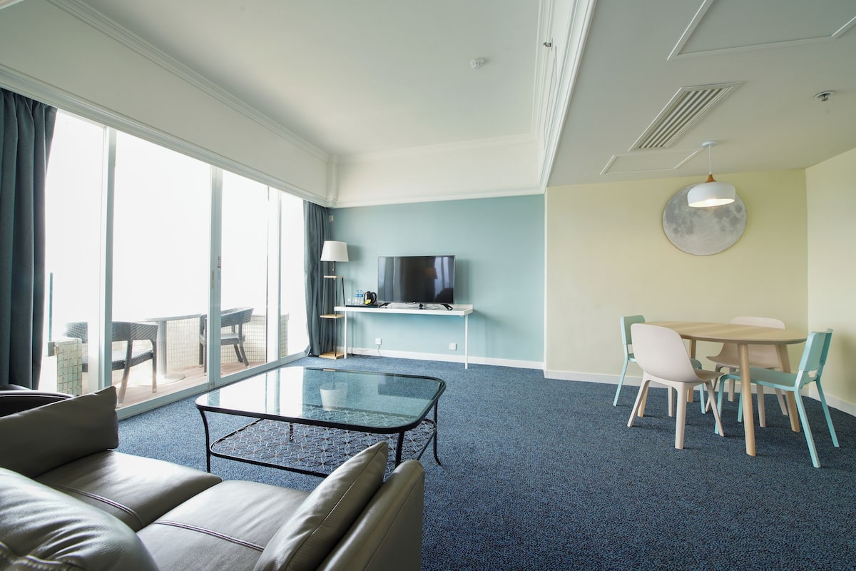 Quarto de hotel moderno com mesa de centro de vidro, sofá de couro, área de jantar com cadeiras, televisão e varanda com mobiliário ao ar livre.