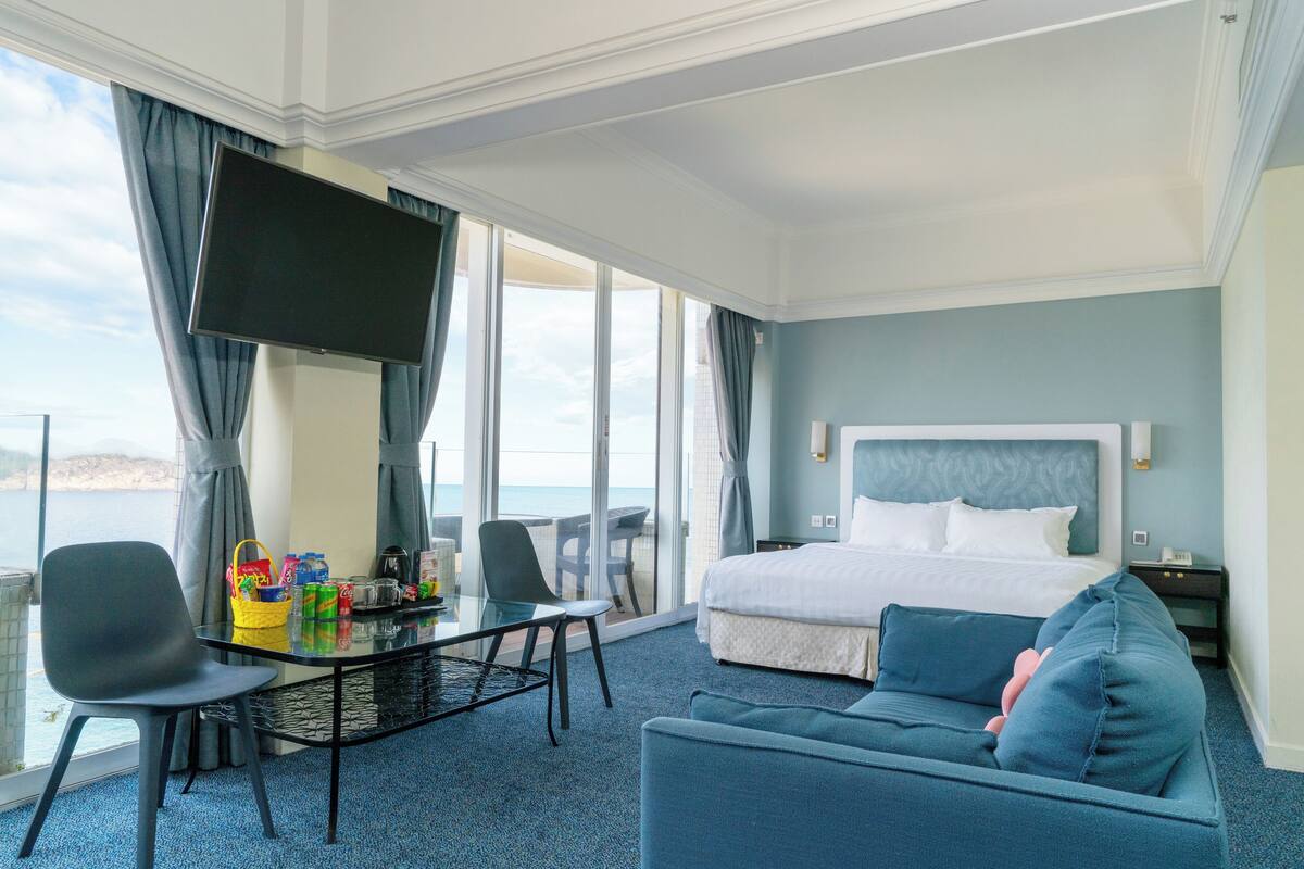 Quarto de hotel com cama, sofá, televisão, mesa com lanches e vista para o mar.