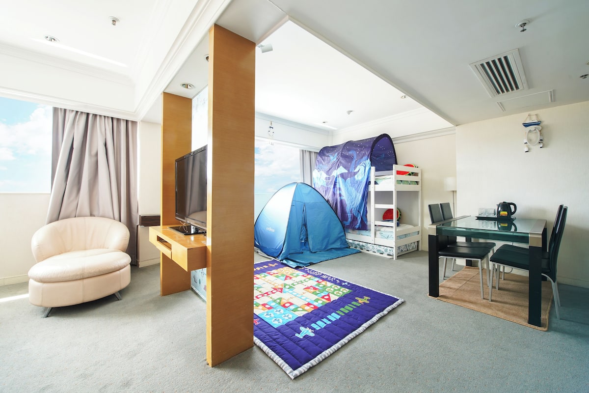 Quarto de hotel moderno com cama, televisão, escrivaninha com cadeira, uma tenda e vista do céu.