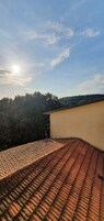 Terraço no telhado com telhas de terracota, céu claro e vista distante de árvores e colinas.