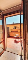 Visão através de uma porta de vidro para uma varanda com piso de azulejos, uma mesa pequena e cadeiras.