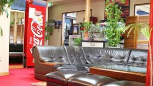 Uma área de estar com sofá de couro, pufes e plantas dentro de casa, além de um anúncio da Coca-Cola.