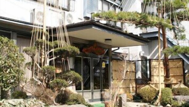 Um edifício japonês tradicional com entrada coberta, rodeado por bonsais bem cuidados e um caminho de pedra.