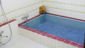 Uma banheira pequena e retangular, com interior azul e exterior em azulejos vermelhos, ao lado de uma parede branca com azulejos estampados em verde.