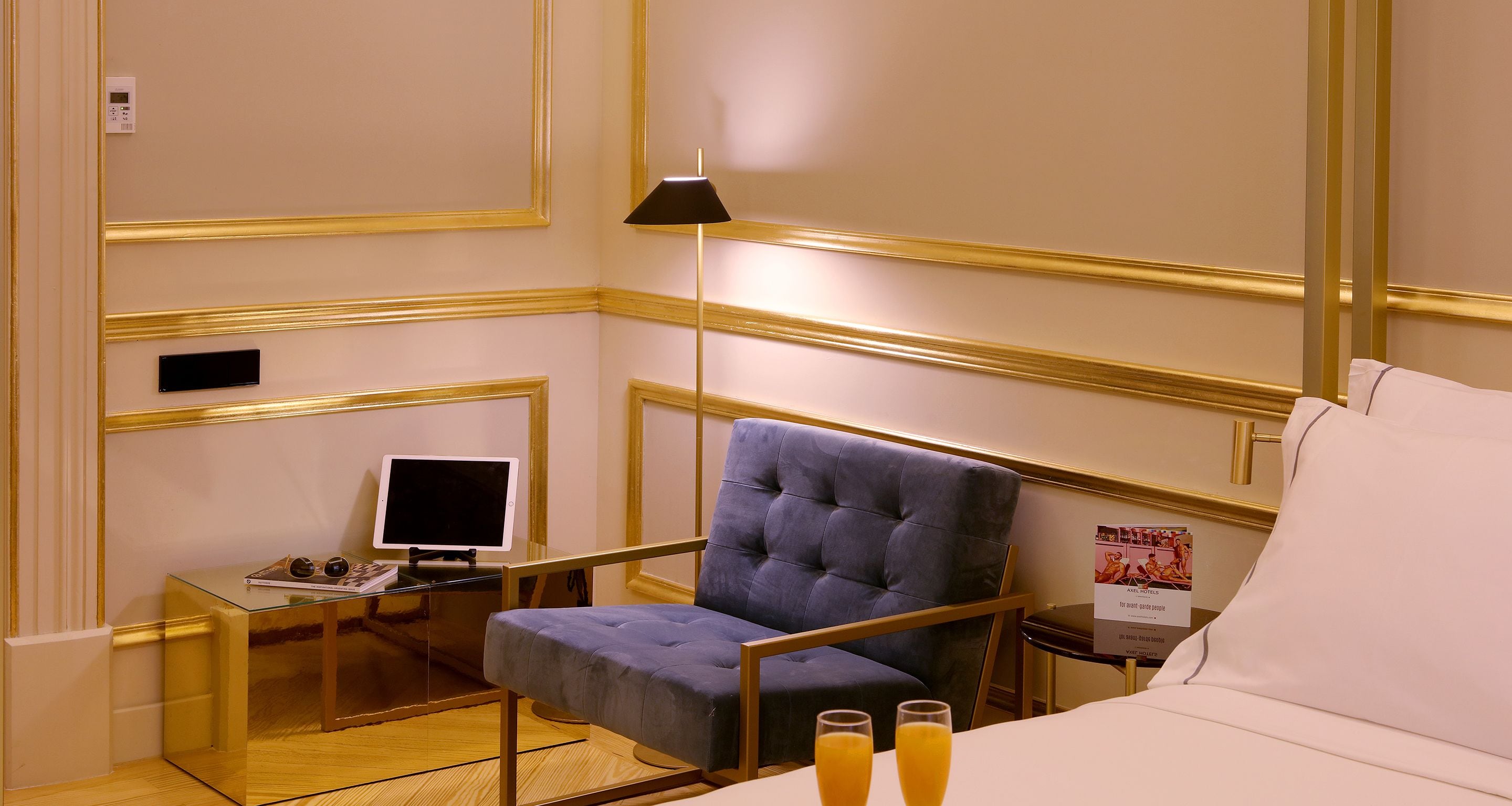 Quarto de hotel com cama, poltrona azul, escrivaninha com computador e pequena mesa com abajur.