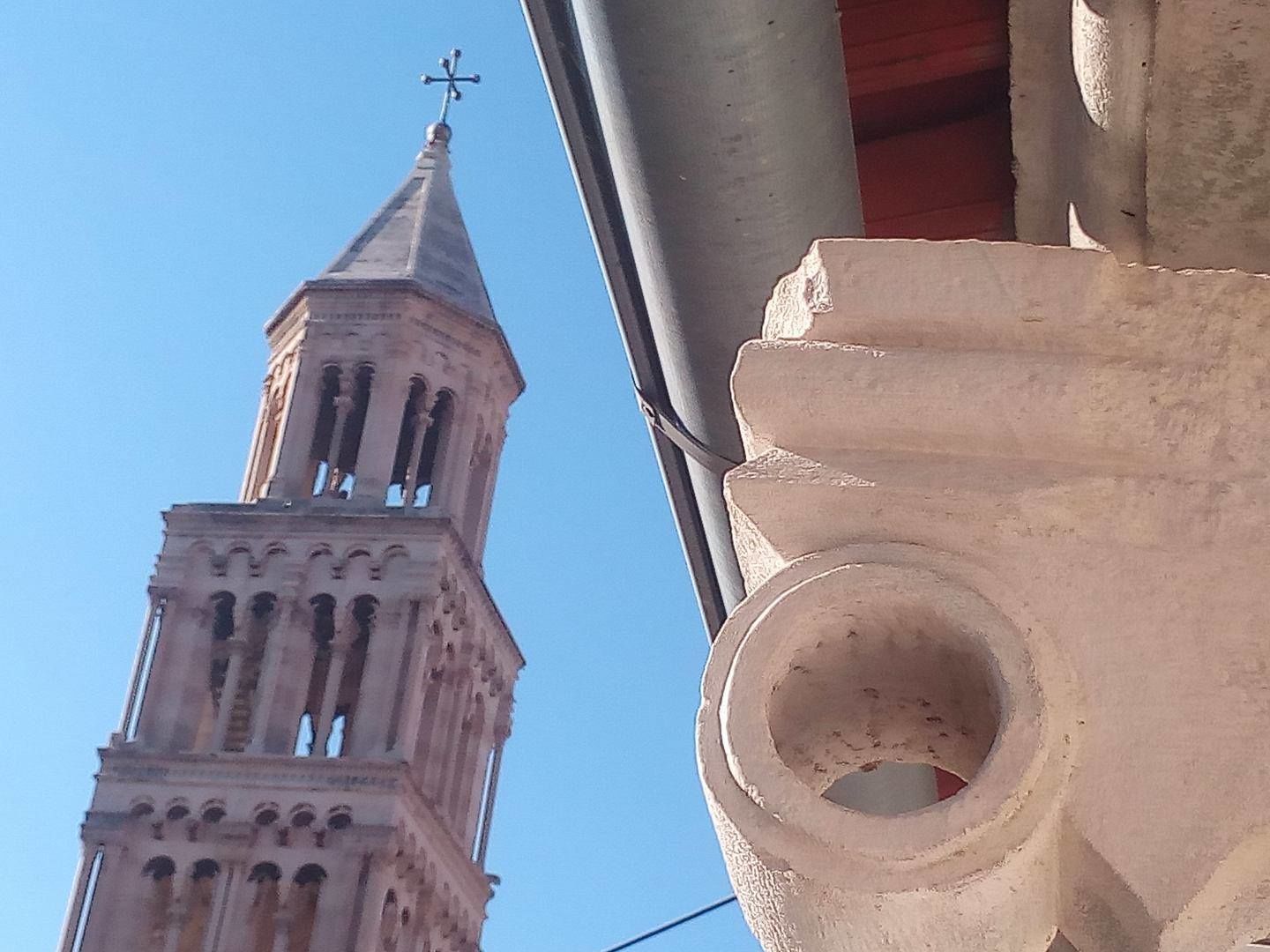 Detalhe do canto de um edifício, com um bico de vento e parte de uma torre com janelas em arco.