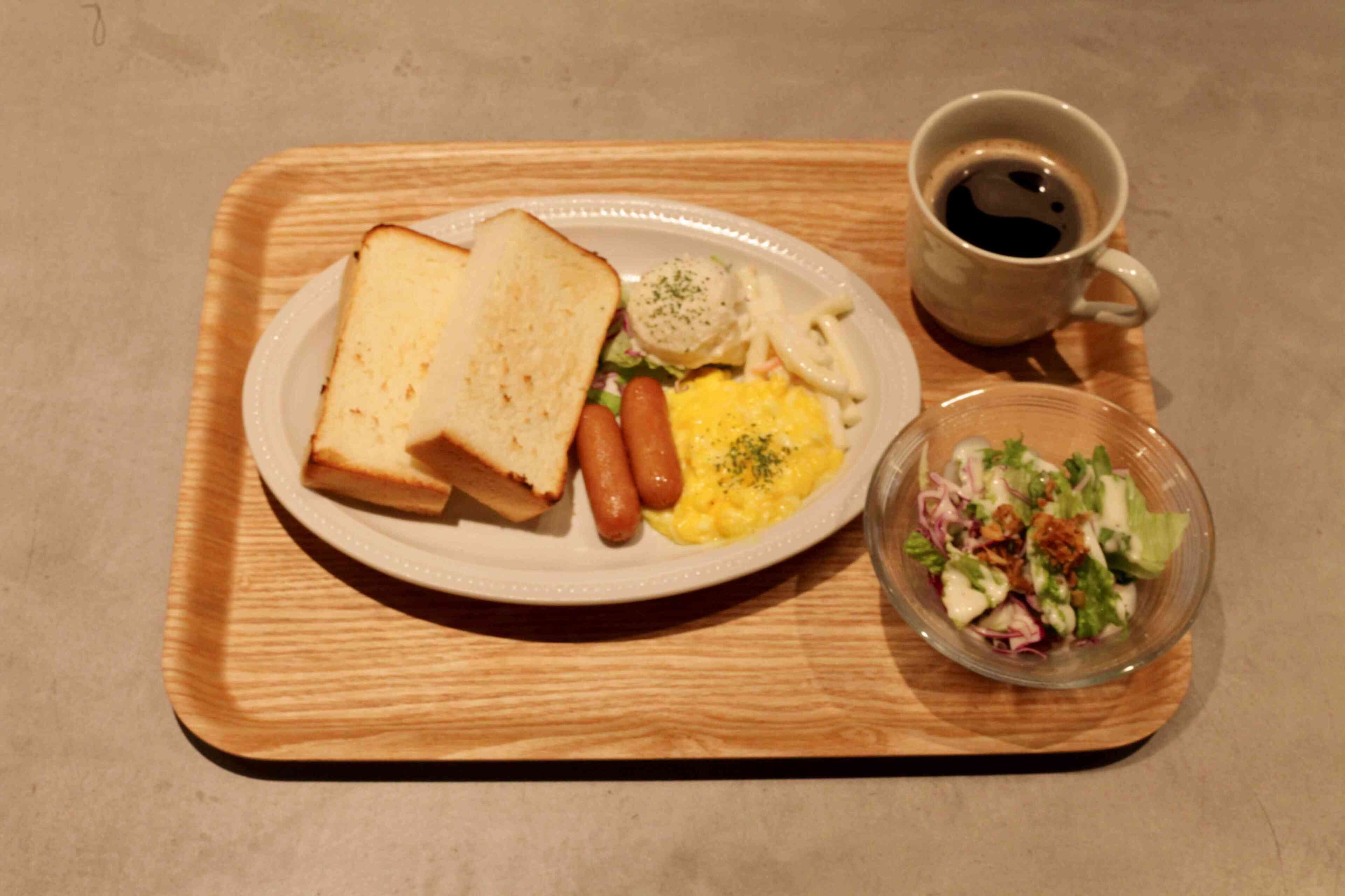 Uma bandeja com torradas, linguiça, ovos mexidos, uma salada pequena e uma xícara de café.