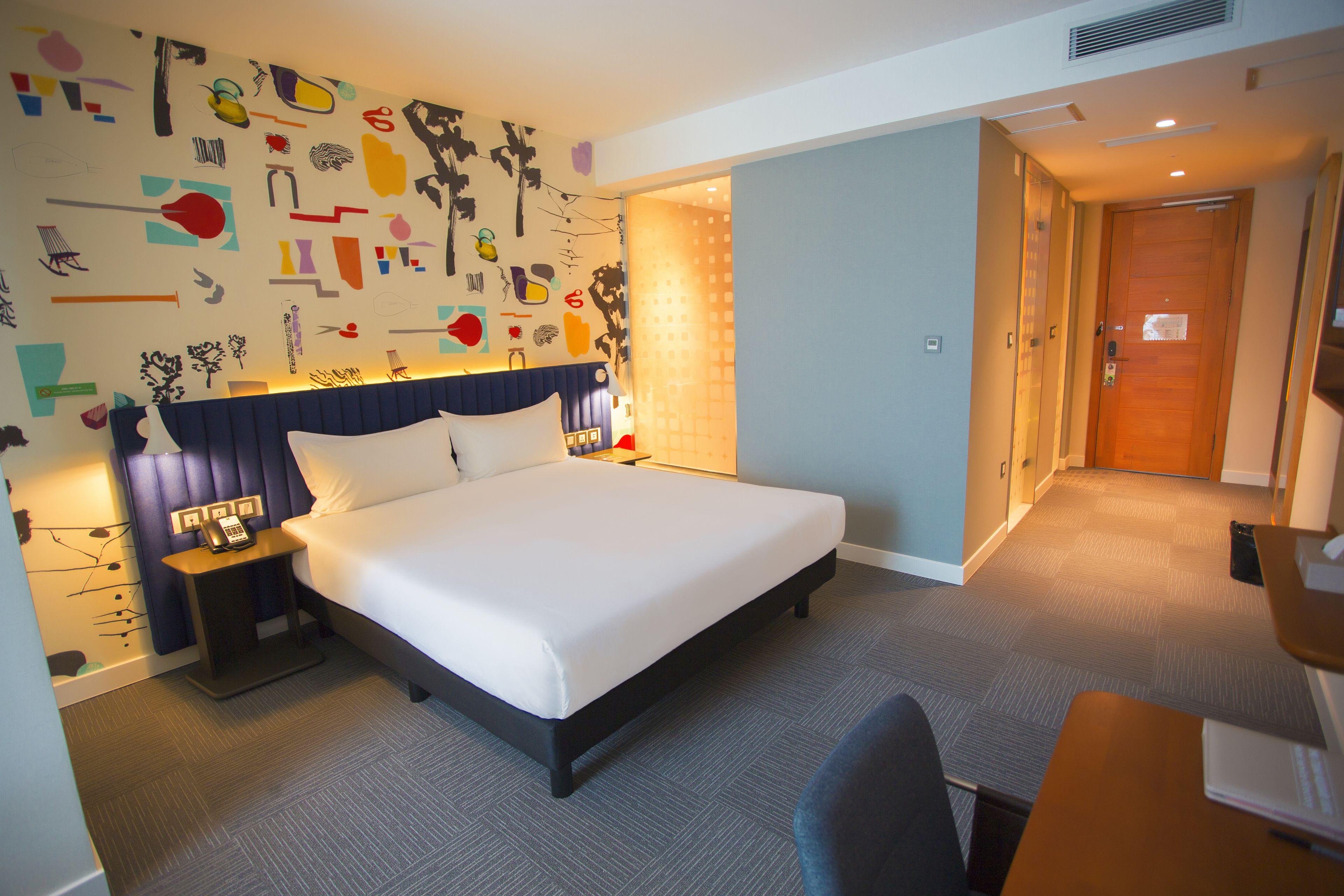 Quarto de hotel com uma cama grande, uma escrivaninha e uma cadeira. Há um telefone na mesinha de cabeceira e uma luminária na escrivaninha.