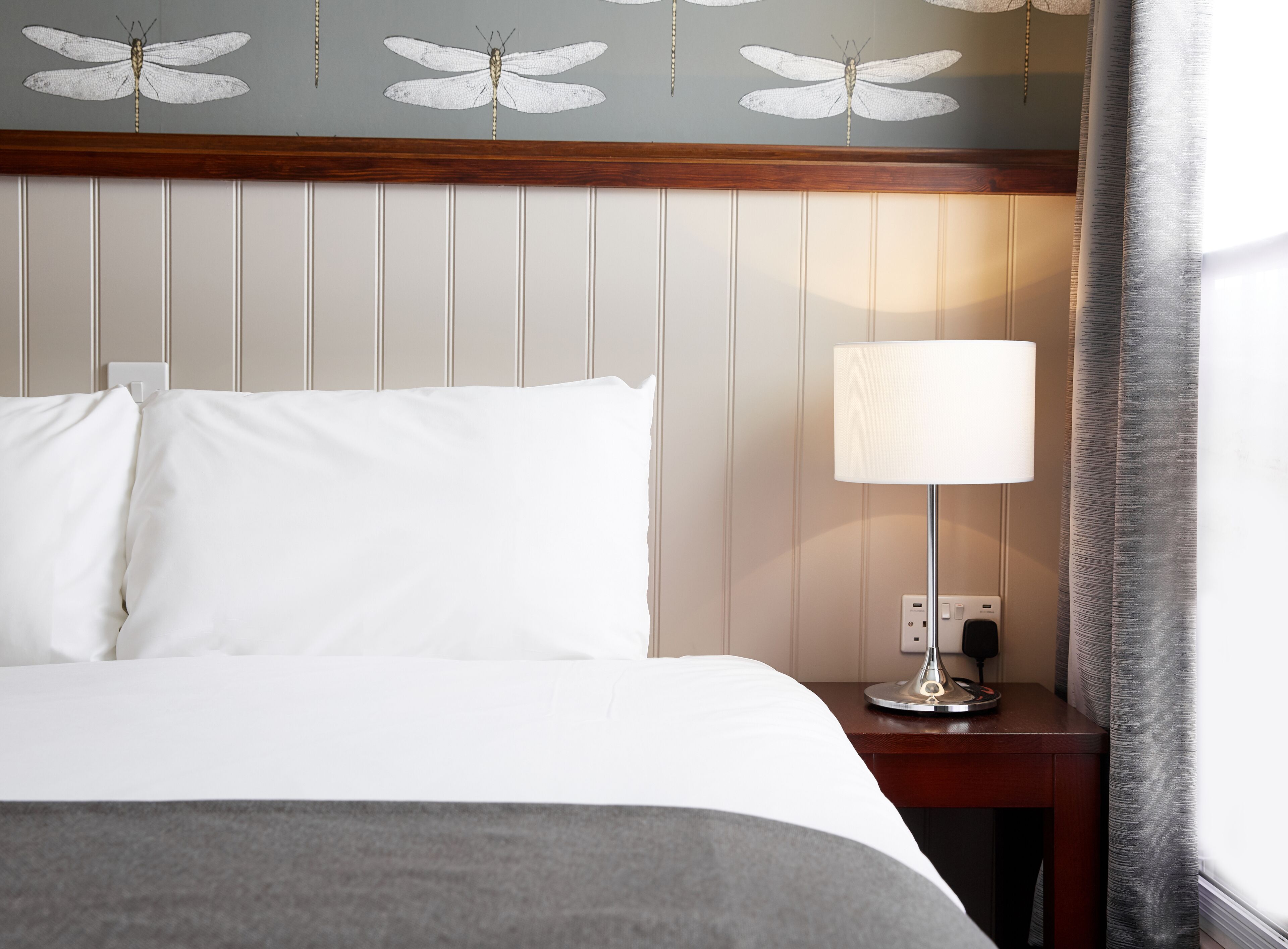 Quarto de hotel com uma cama, uma mesa de cabeceira com abajur e uma parede decorada com adesivos de borboletas.
