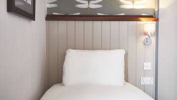 Cama bem arrumada com travesseiro branco e manta cinza, luminária fixada na parede e quadro emoldurado na parede.