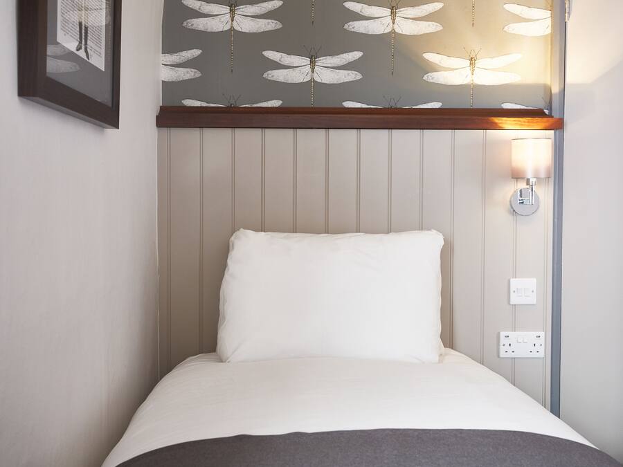 Cama bem arrumada com travesseiro branco e manta cinza, luminária fixada na parede e quadro emoldurado na parede.