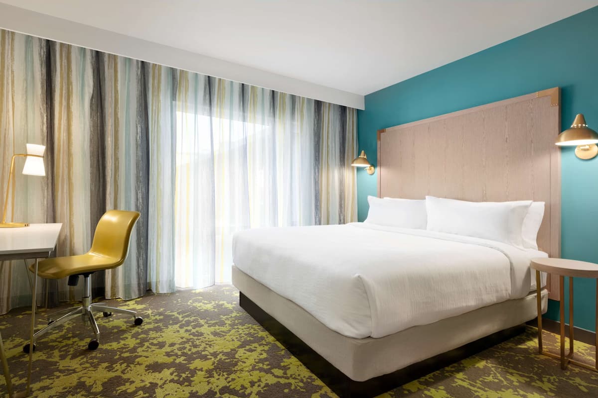 Quarto de hotel com uma cama grande, uma escrivaninha com cadeira e uma janela com cortinas.