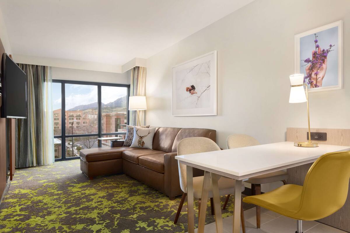 Quarto de hotel moderno com uma janela grande, um sofá, uma mesa de jantar e uma televisão.