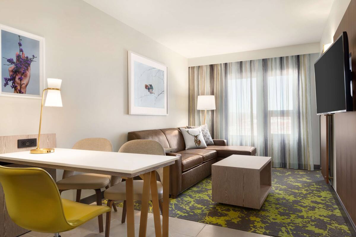 Quarto de hotel moderno com uma mesa de jantar, cadeiras amarelas, um sofá de couro marrom, uma mesa de centro de madeira, uma TV de tela plana e uma janela grande com cortinas.