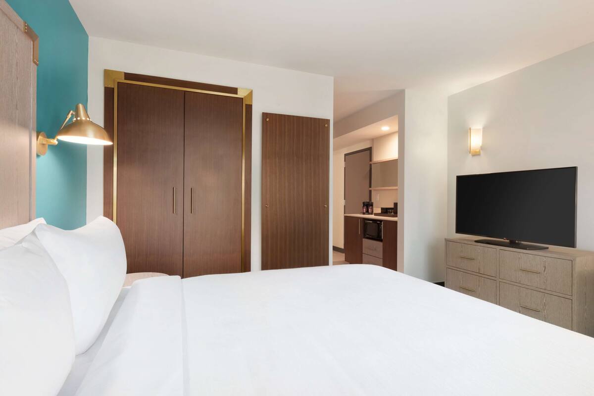 Quarto de hotel com cama, televisão, guarda-roupa e cozinha compacta.