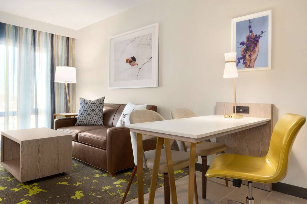 Quarto de hotel moderno com um sofá de couro marrom, uma mesa branca, uma cadeira amarela e uma grande janela com cortinas.