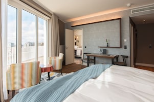 Quarto de hotel com uma cama grande, uma escrivaninha, duas cadeiras, uma mesa pequena e vista da cidade através de grandes janelas.