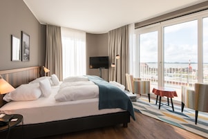 Quarto de hotel com cama grande, televisão, escrivaninha com candeeiro e varanda com cadeiras.