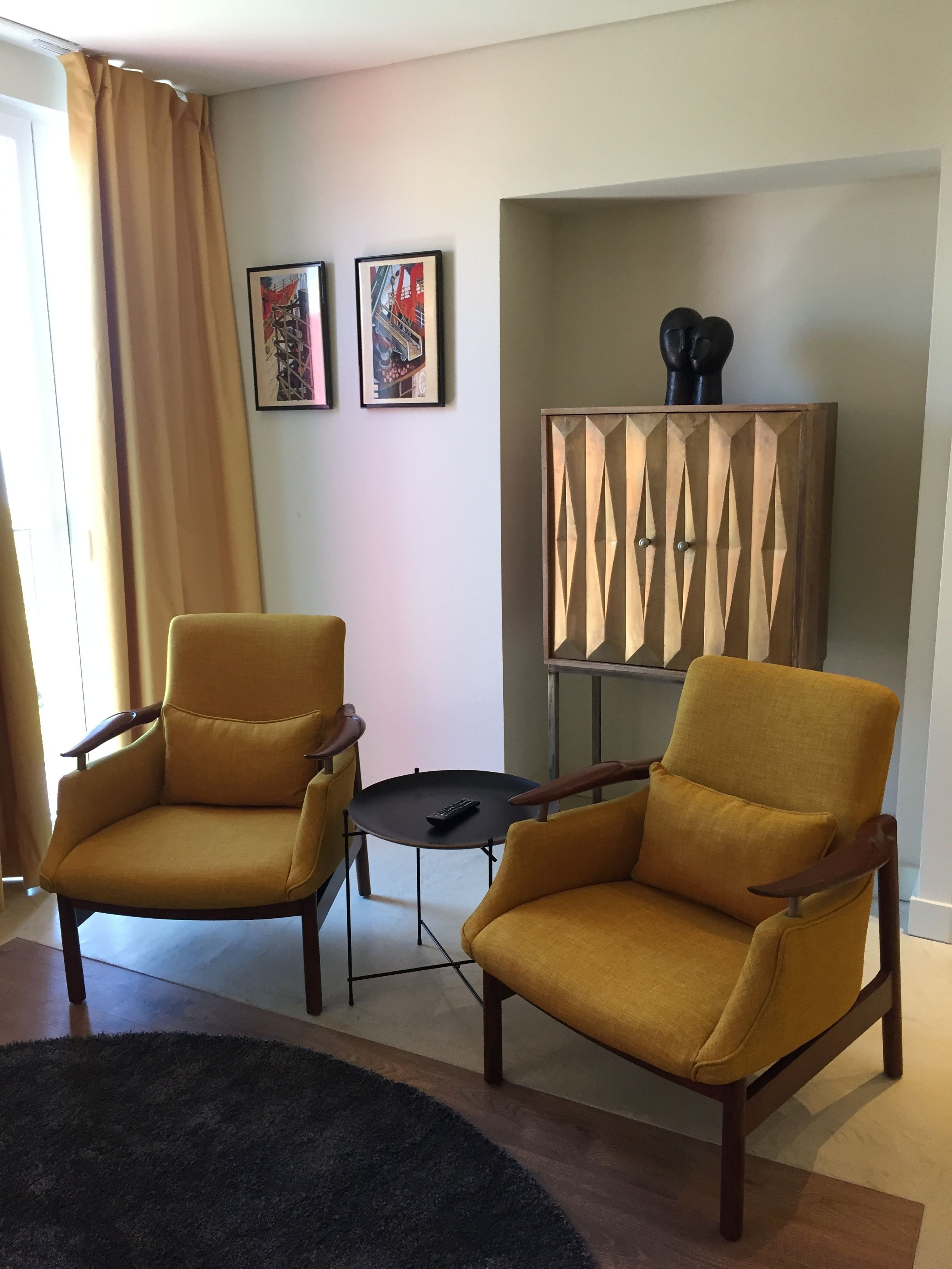 Sala de estar com dois poltronas cor mostarda, uma mesinha redonda e um tapete preto. Há duas molduras na parede e um armário com padrões geométricos.