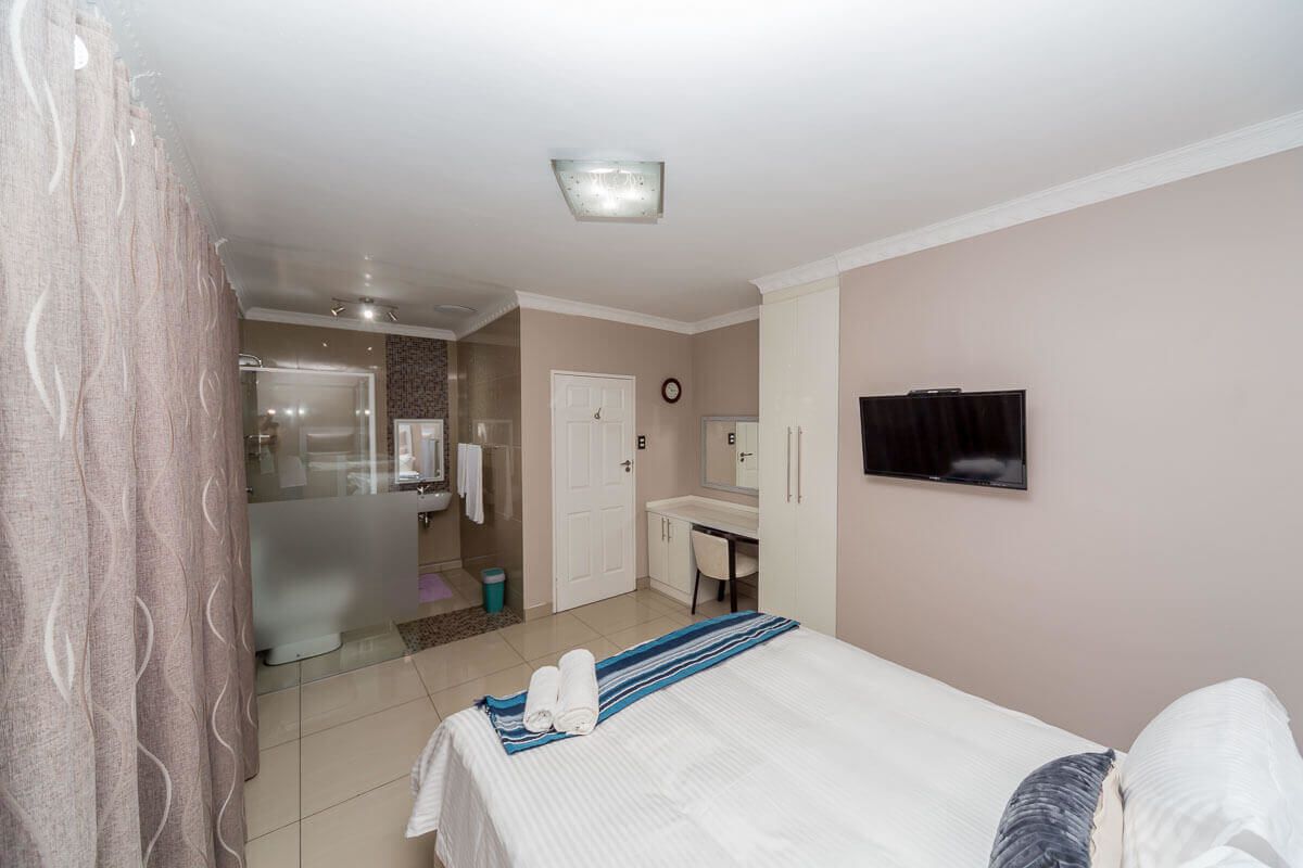 Quarto de hotel com cama, escrivaninha, espelho, televisão e banheiro com chuveiro e vaso sanitário.