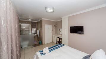 Quarto de hotel com cama, escrivaninha, espelho, televisão e banheiro com chuveiro e vaso sanitário.
