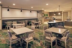 Um saguão de hotel com várias áreas de estar, incluindo assentos individuais e mesas, uma cozinha e um balcão de recepção.