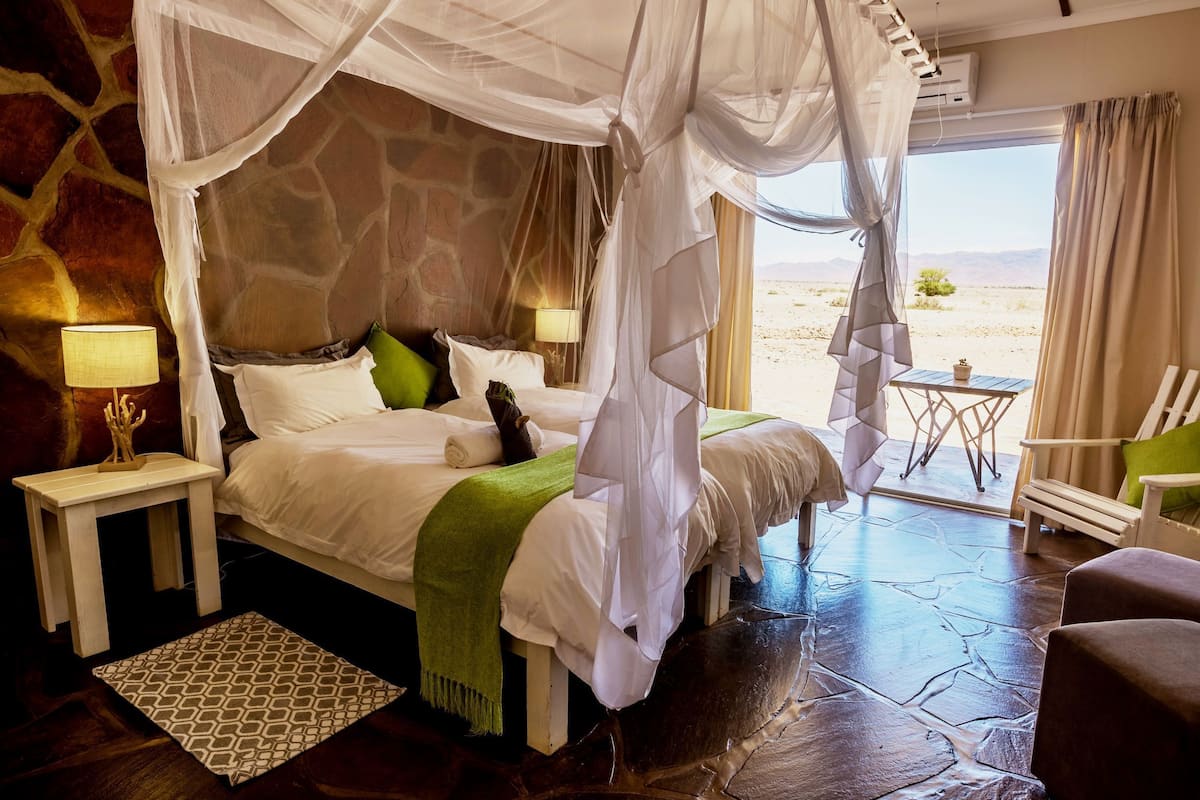 Quarto com parede de pedra, cama com roupa de cama branca e manta verde, mesa de cabeceira com abajur, e vista para o deserto através de uma janela grande.