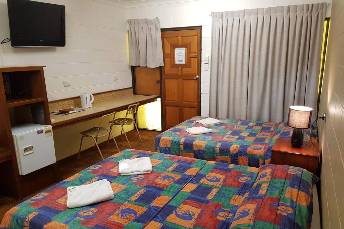 Quarto com duas camas, uma escrivaninha, uma cadeira, uma TV e uma porta.
