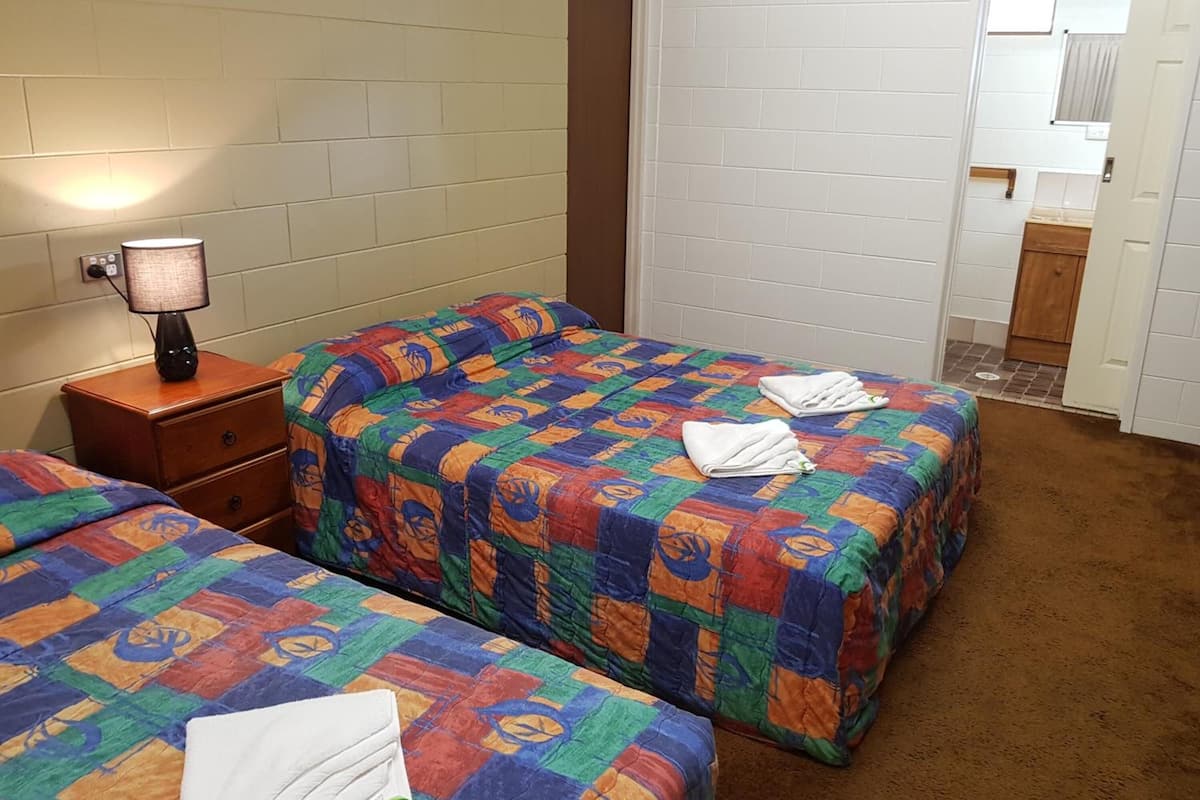 Quarto de hotel com duas camas, uma mesa de cabeceira, uma luminária e um banheiro visível através de uma porta aberta.