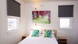 Quarto com uma cama, duas mesas de cabeceira, um quadro abstrato na parede e duas janelas com persianas.