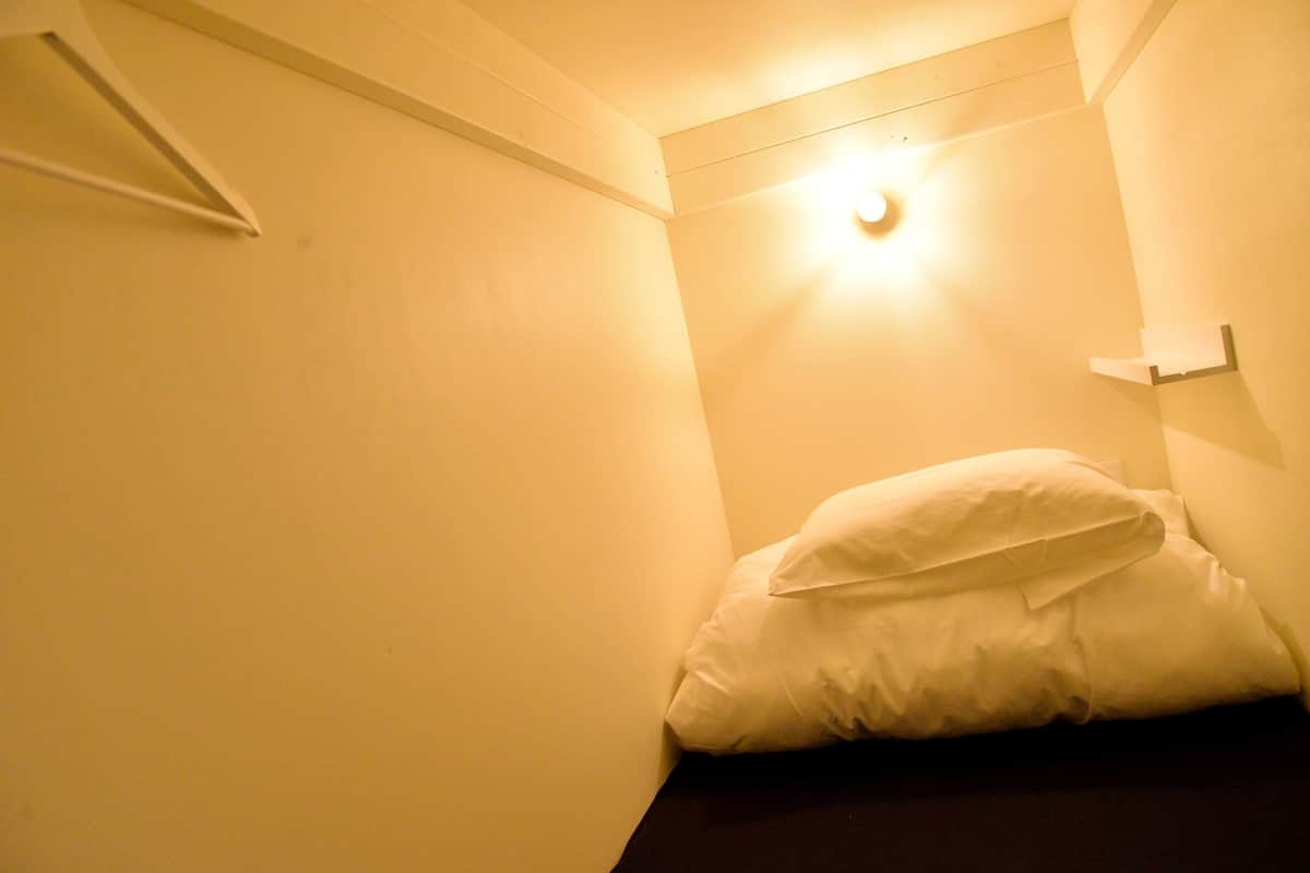 Canto de um quarto com uma cama, uma parede branca e um luminário.