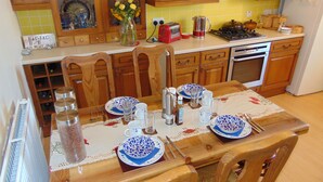 Uma área de refeições com cadeiras de madeira e uma mesa posta para quatro, com pratos estampados em azul e branco e toalha de renda.
