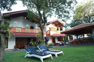 Um edifício de dois andares com fachada vermelha e branca, um espaço coberto com varanda e um gramado com cadeiras de sol.